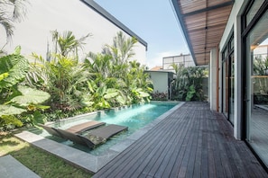 Uma área de piscina moderna ao ar livre, com um deck de madeira, rodeada por plantas tropicais exuberantes e uma piscina azul cristalina.