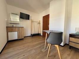Quarto pequeno e limpo com piso de madeira, uma escrivaninha com cadeira, televisão montada na parede, porta de madeira e um armário pequeno.
