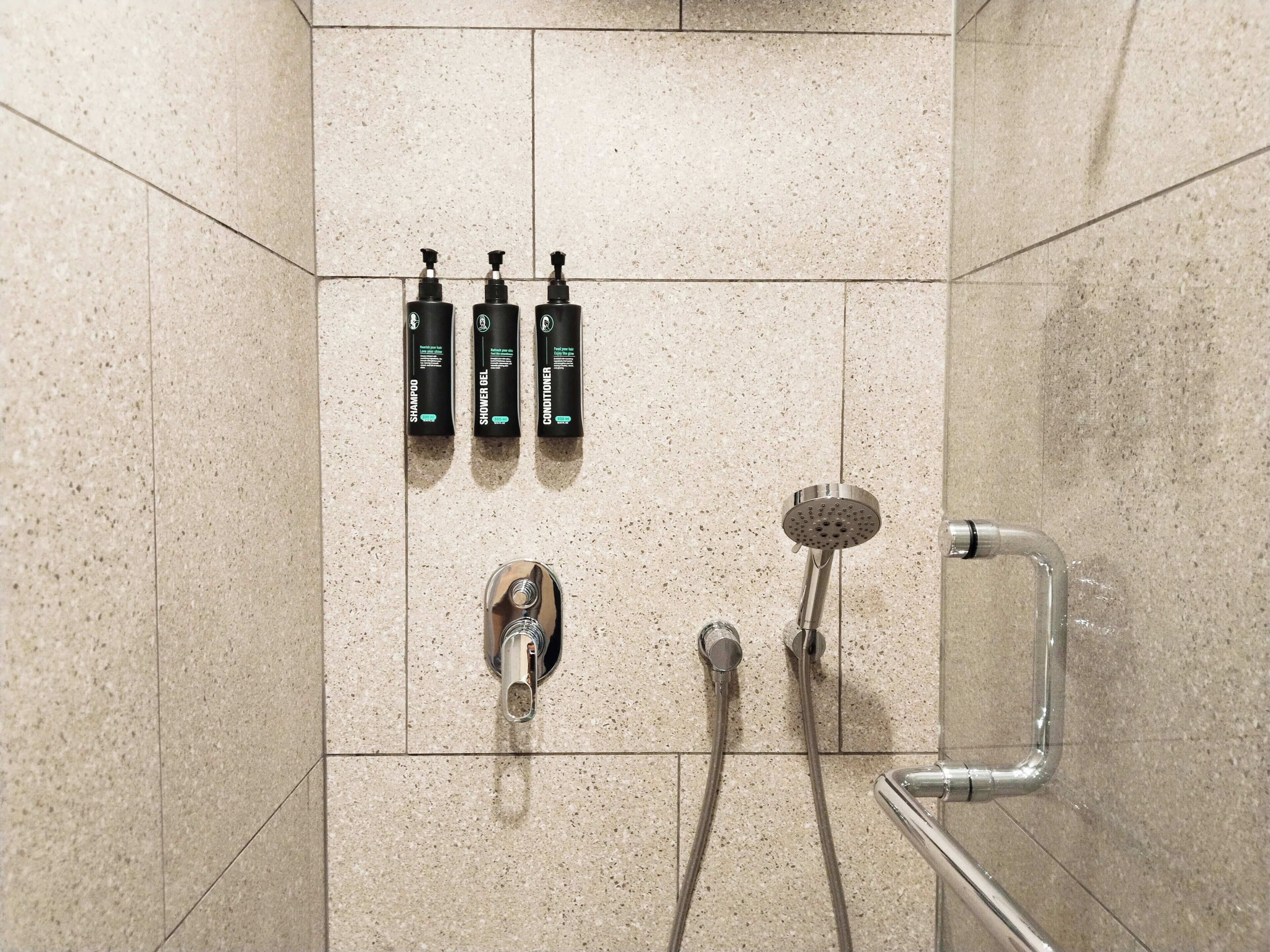 Área de duche com três garrafas de champô, condicionador e gel de banho, um chuveiro manual e uma torneira fixada na parede.