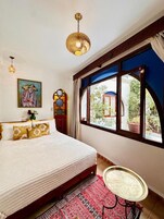 Um quarto com cama, luminária pendente, janela com cortinas e uma mesinha.