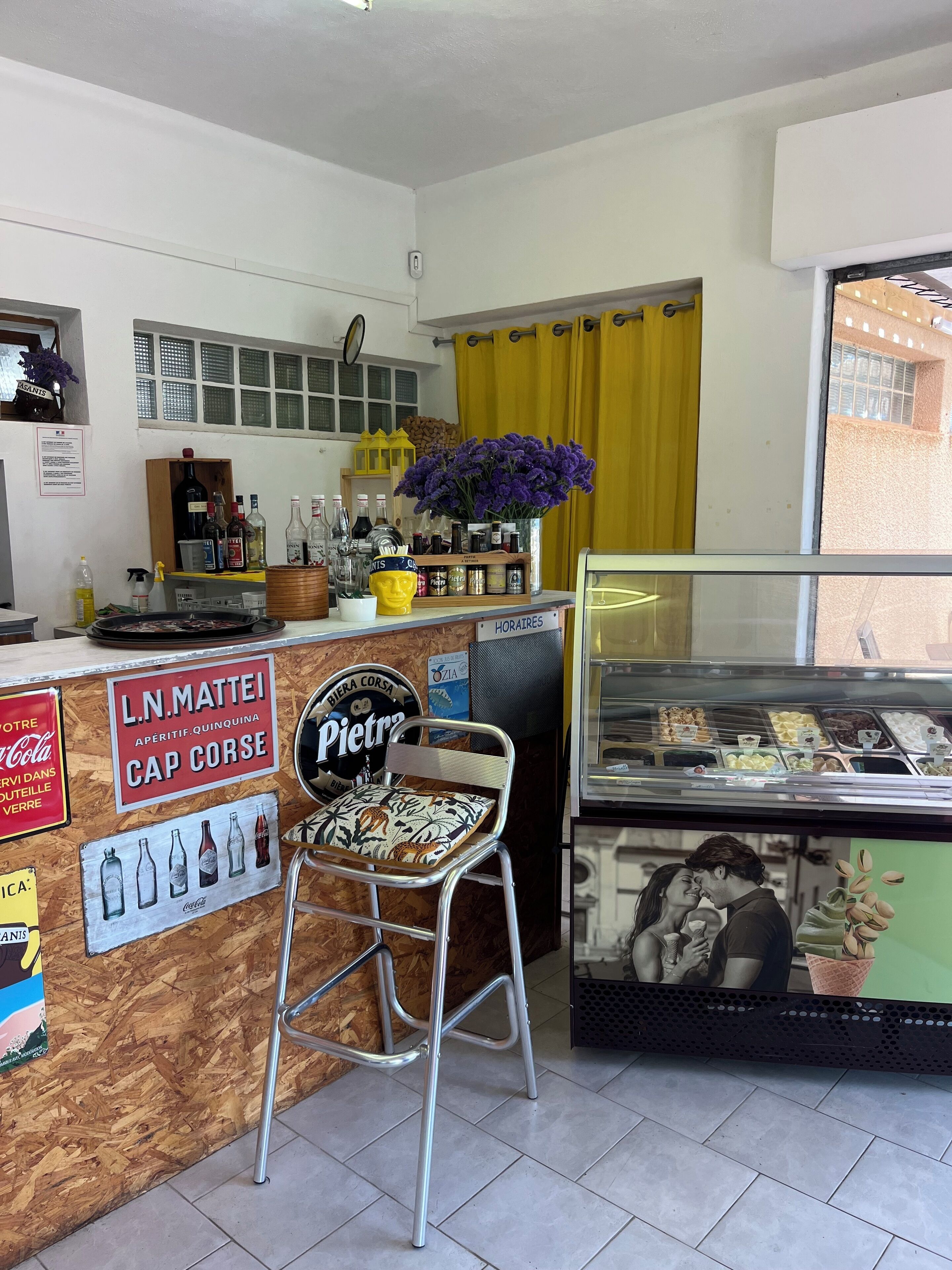 Interior de café com balcão de madeira, cortina amarela, vitrine de vidro e uma cadeira com almofada estampada.
