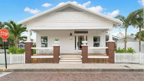 Uma casa branca de dois andares com varanda frontal, colunas de tijolo e cerca branca de piquete.