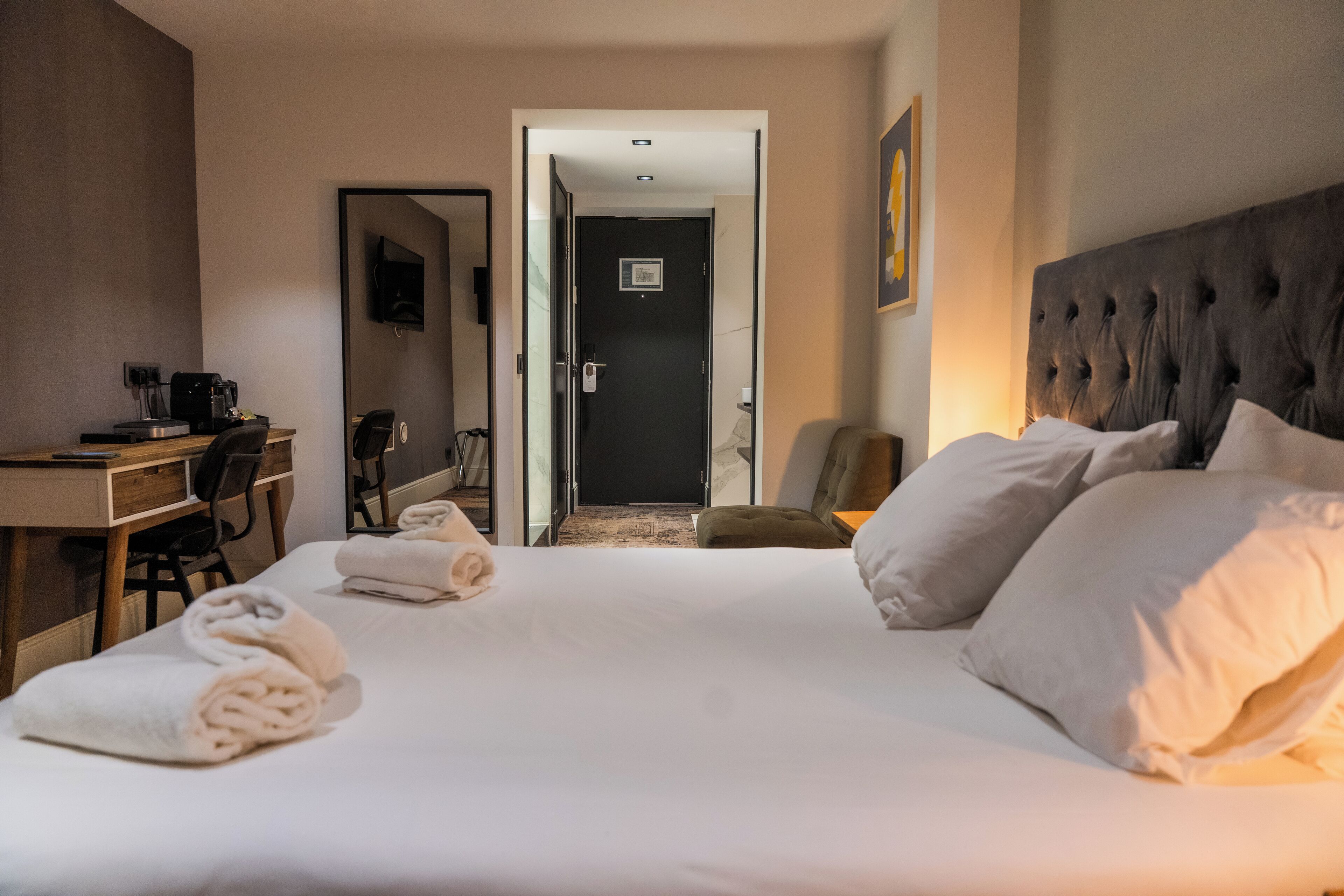 Quarto de hotel com cama, escrivaninha, cadeira, televisão e porta que dá para outro cômodo.