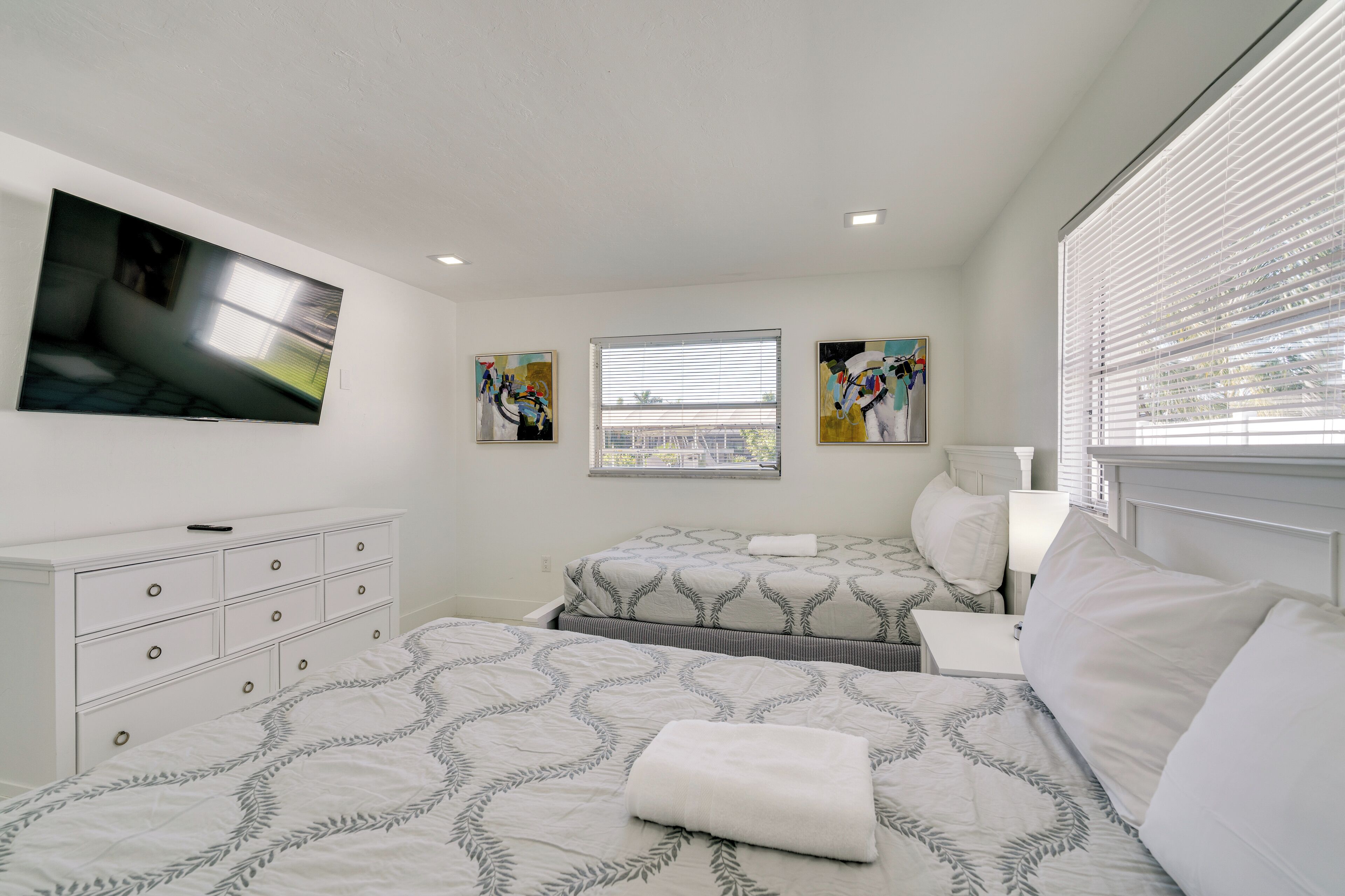 Quarto com duas camas, televisão, obras de arte e janela com persianas.