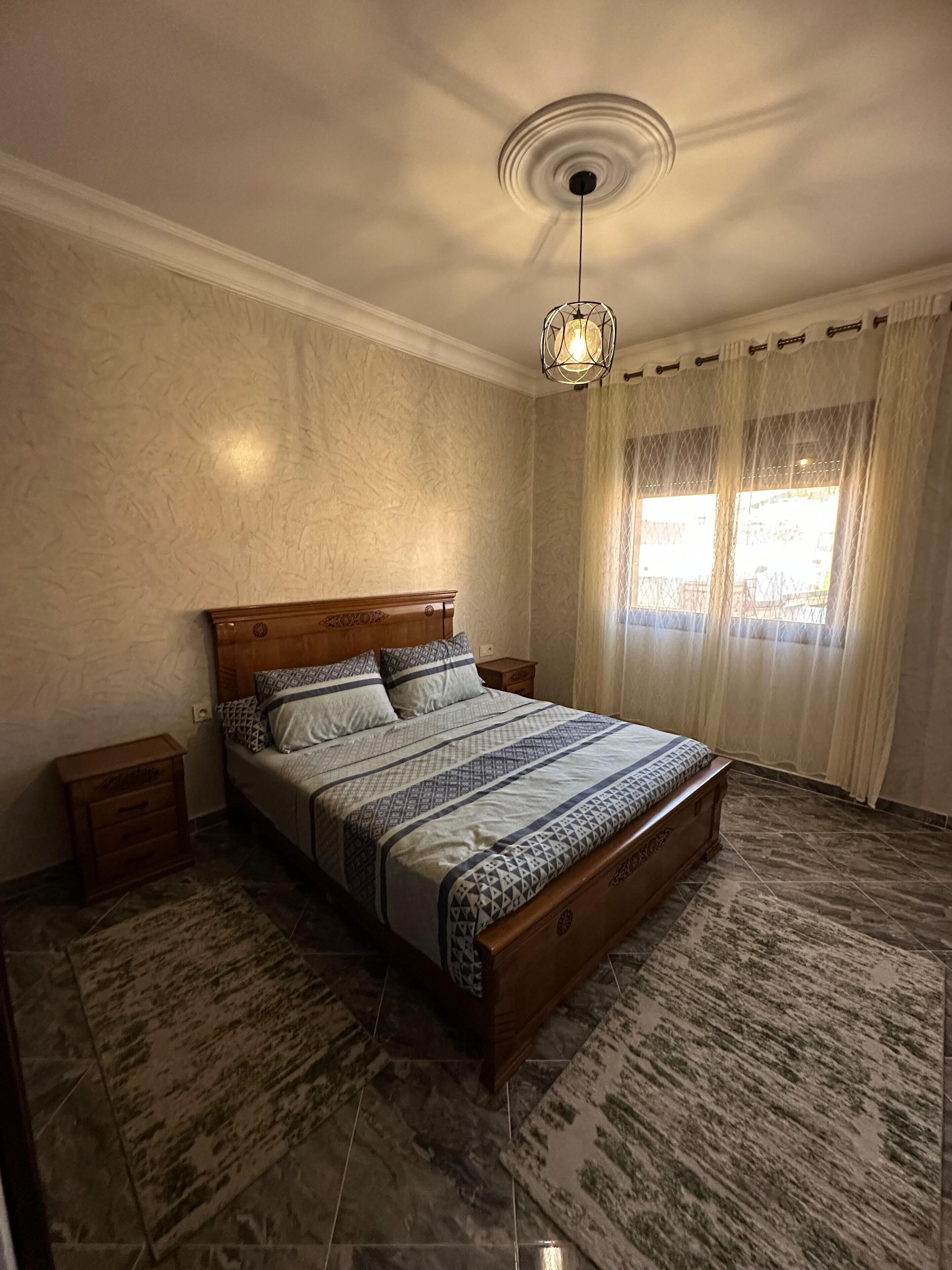 Quarto com cama de madeira, roupa de cama estampada, mesa de cabeceira, luminária no teto e vista pela janela com cortinas.