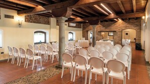 Uma sala de conferências com cadeiras brancas dispostas em fileiras, vigas de madeira no teto e paredes de tijolo.