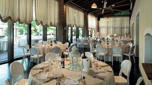 Uma sala de banquetes com mesas redondas preparadas para refeições, cadeiras brancas e uma grande janela com cortinas.