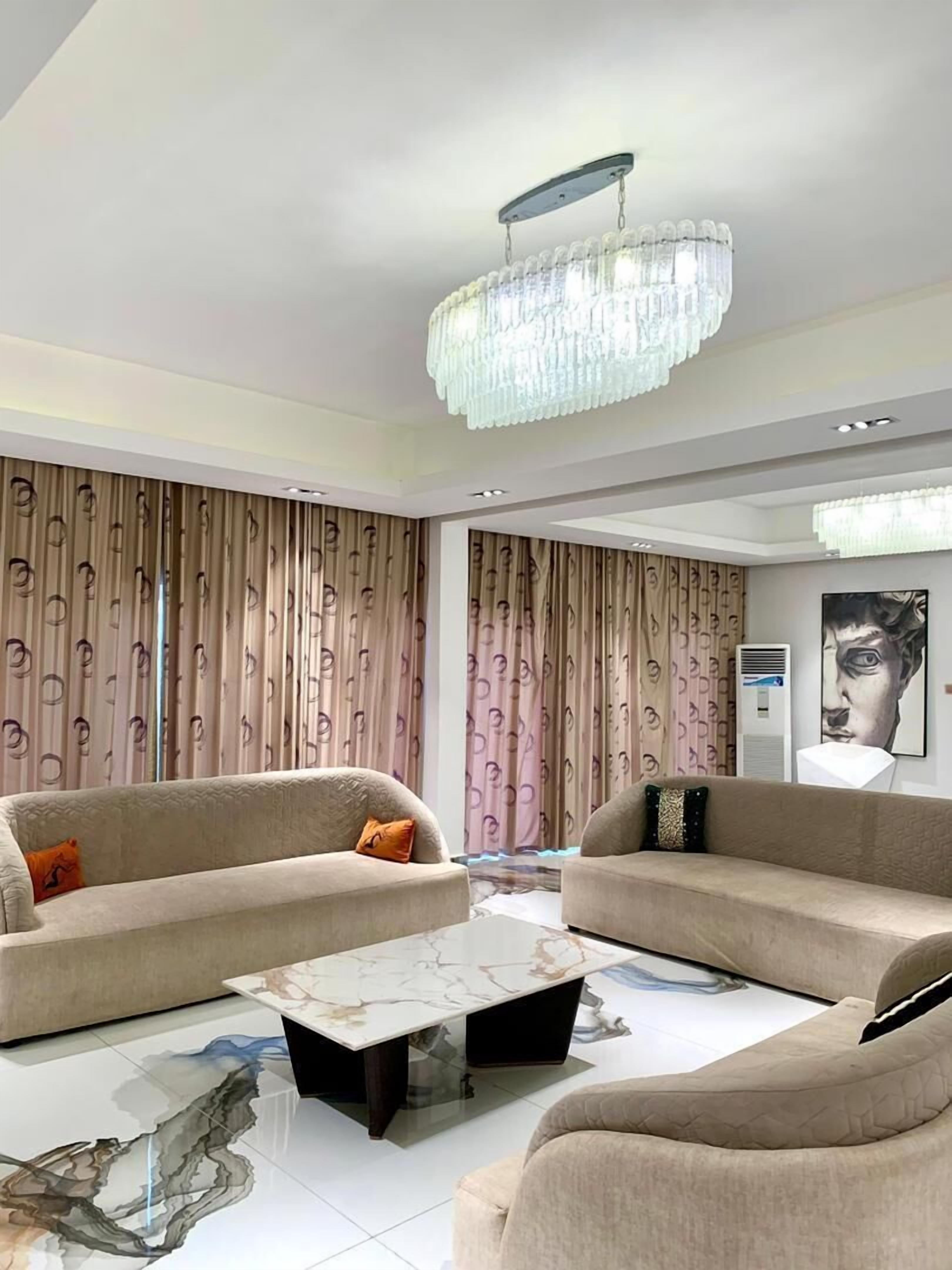 Uma sala de estar moderna com dois sofás, uma mesa de centro, um candeeiro de lustre e cortinas estampadas.