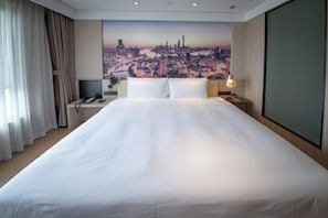 Quarto de hotel com uma cama grande, um mural da paisagem urbana, uma mesa de cabeceira com abajur e uma janela com cortinas.