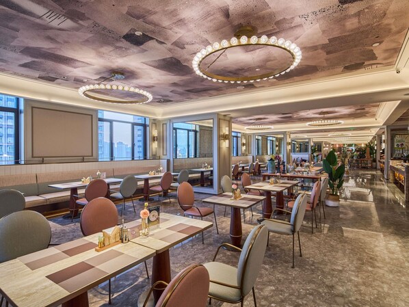 Ampla sala de jantar de hotel com várias mesas e cadeiras, grandes janelas e um design de teto moderno.