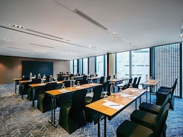 Uma sala de conferências com várias mesas e cadeiras, janelas amplas e piso estampado.