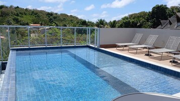Piscina no terraço com água azul cristalina, cercada por guarda-corpo de vidro e espreguiçadeiras brancas.