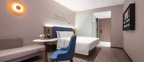 Quarto de hotel moderno com uma cama, uma cadeira azul, uma mesa redonda pequena e uma luminária fixada na parede.