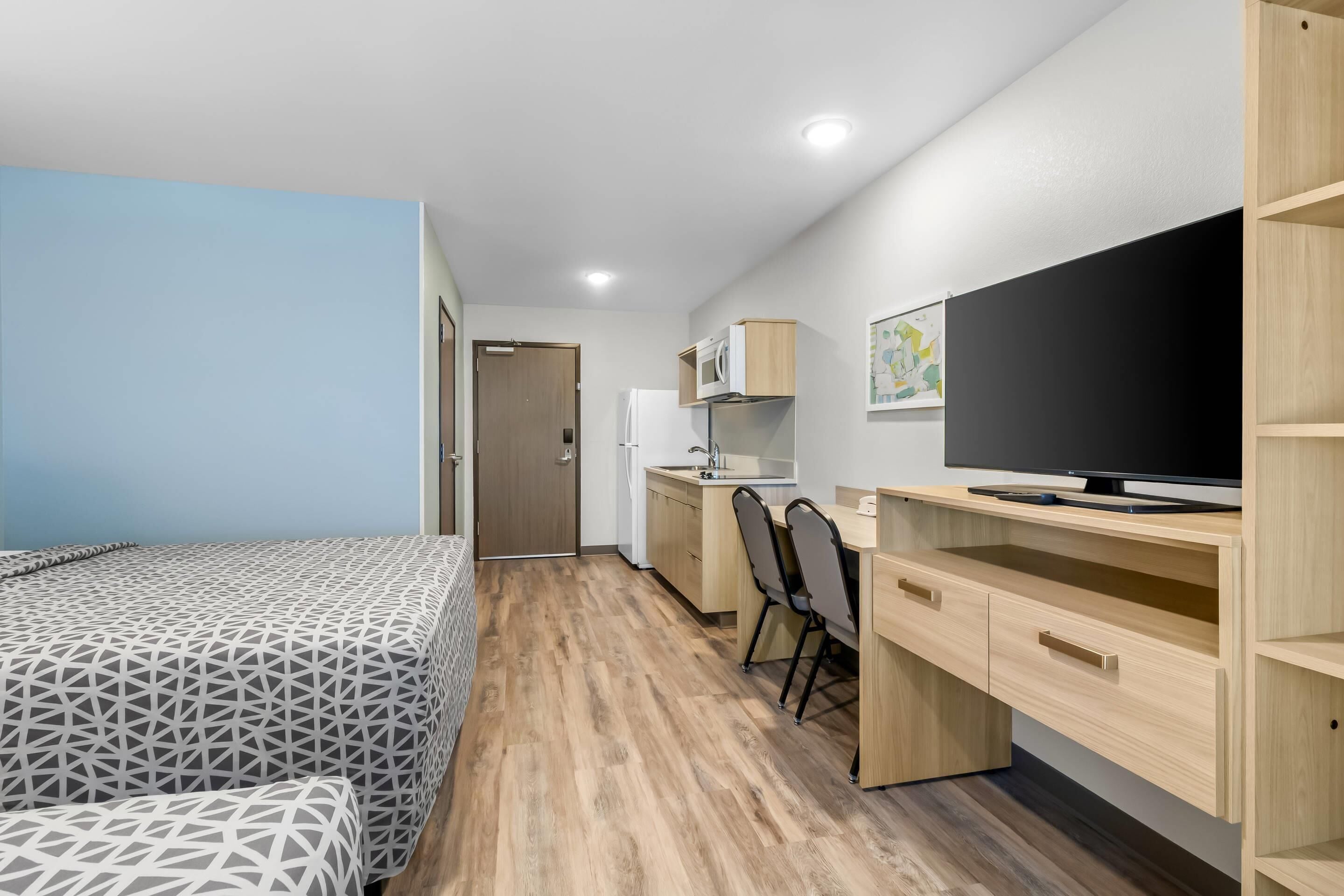 Quarto compacto com cama, escrivaninha, cadeira, televisão e micro-ondas.