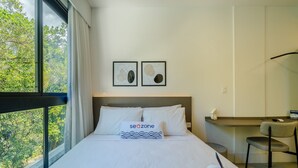 Quarto de hotel com uma cama, escrivaninha, cadeira e janela com vista para árvores.