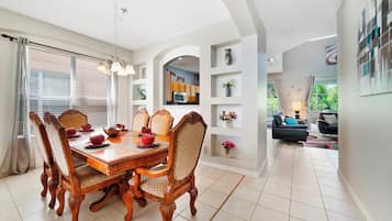 Uma área de refeições com uma mesa e cadeiras de madeira, uma cozinha ao fundo e uma sala de estar com um sofá e abajur.