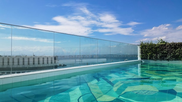 Piscina infinita no terraço com barreira de vidro, céu azul límpido e vista distante de uma ponte.