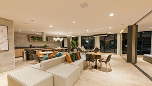 Uma sala de estar moderna com um sofá seccional, uma área de jantar com mesa redonda e uma cozinha ao fundo.