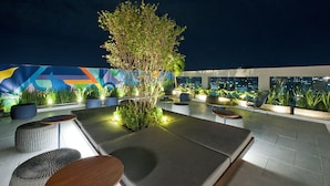 Terraço no telhado com áreas de estar, vasos de plantas e vista para a cidade à noite.
