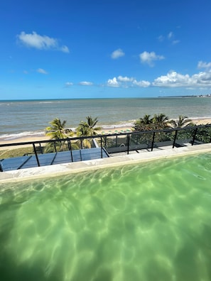 Visão clara de uma piscina com barreira de vidro, uma praia com palmeiras e uma linha costeira ao longe.