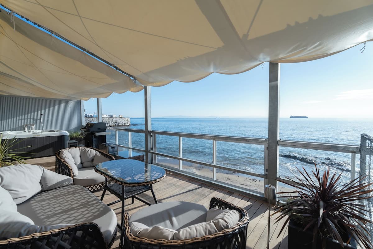 Uma área de estar coberta ao ar livre, com móveis de vime, uma mesa e vista para o mar.