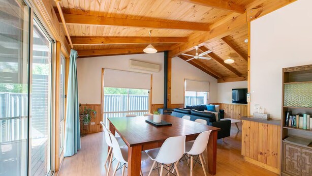 Um quarto espaçoso com teto de madeira, uma mesa de jantar com cadeiras, um sofá e uma área de cozinha com geladeira e prateleiras.