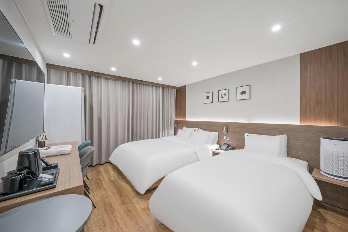 Quarto de hotel com duas camas, uma mesa com cafeteira, televisão e purificador de ar fixado na parede.