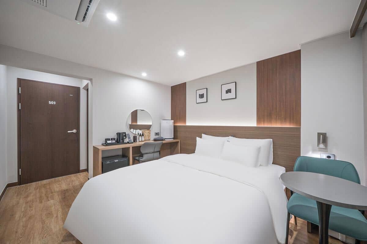 Quarto de hotel com uma cama grande, uma escrivaninha com cadeira, uma mesa pequena, um espelho e uma porta com a inscrição '509'.