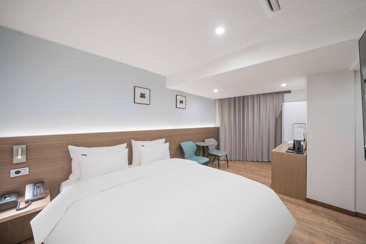 Quarto de hotel moderno com uma cama grande, telefone ao lado da cama, uma pequena escrivaninha com luminária e uma televisão.