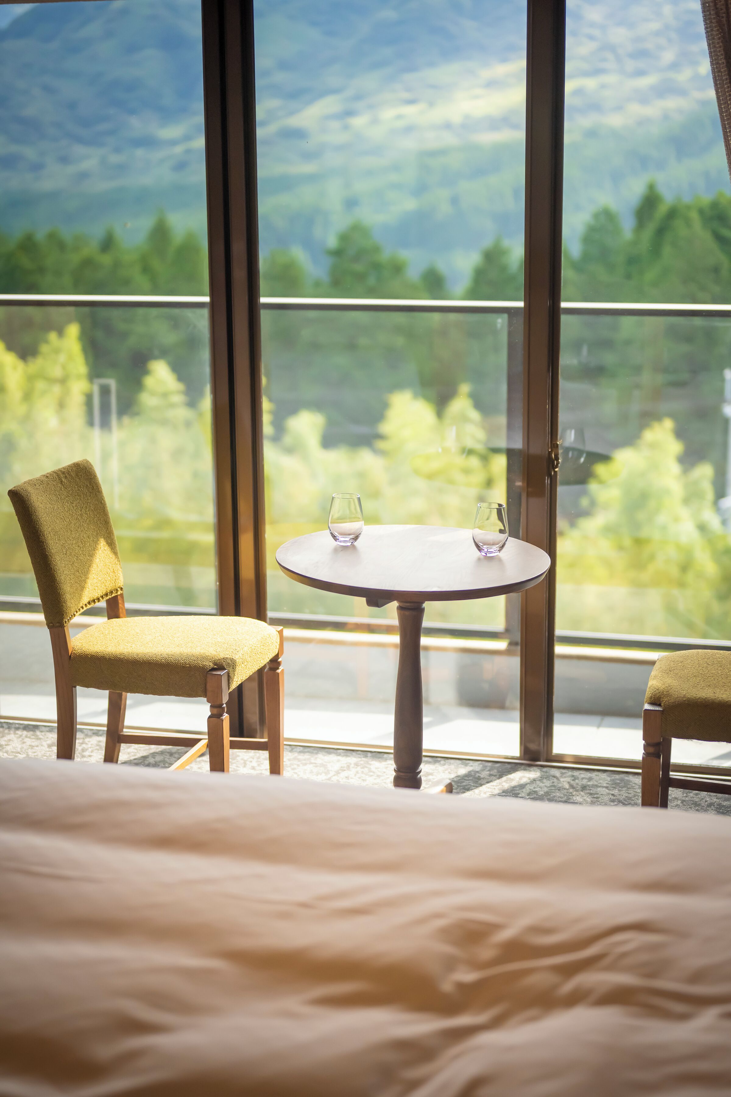 Quarto com uma cama, uma mesa redonda com duas cadeiras e uma janela grande que dá vista para árvores e montanhas.