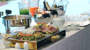 Uma mesa de buffet com diversos alimentos e bebidas, incluindo uma salada, carnes e uma seleção de bebidas em recipiente de vidro.