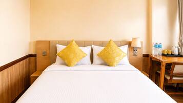 Quarto de hotel com cama, duas almofadas, mesa de cabeceira, luminária e mesa com garrafas de água e cafeteira.