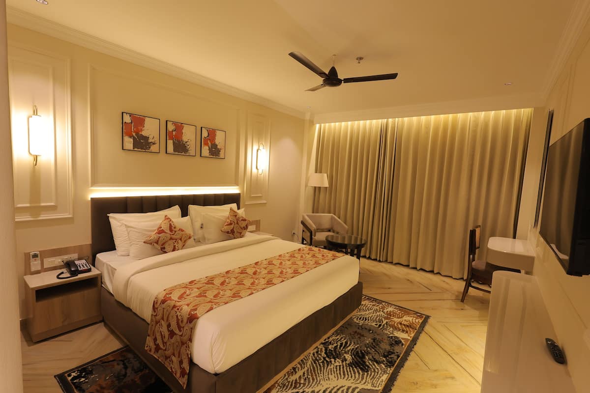 Quarto de hotel com uma cama grande, uma escrivaninha com luminária, uma cadeira, uma televisão e um ventilador de teto.