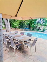 Uma área de refeições ao ar livre com mesa e cadeiras sob um toldo, piscina ao fundo e um cenário natural com árvores e pedras.