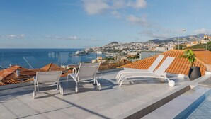 Área de piscina no terraço com cadeiras de lounge brancas e vista para o mar e o horizonte urbano.
