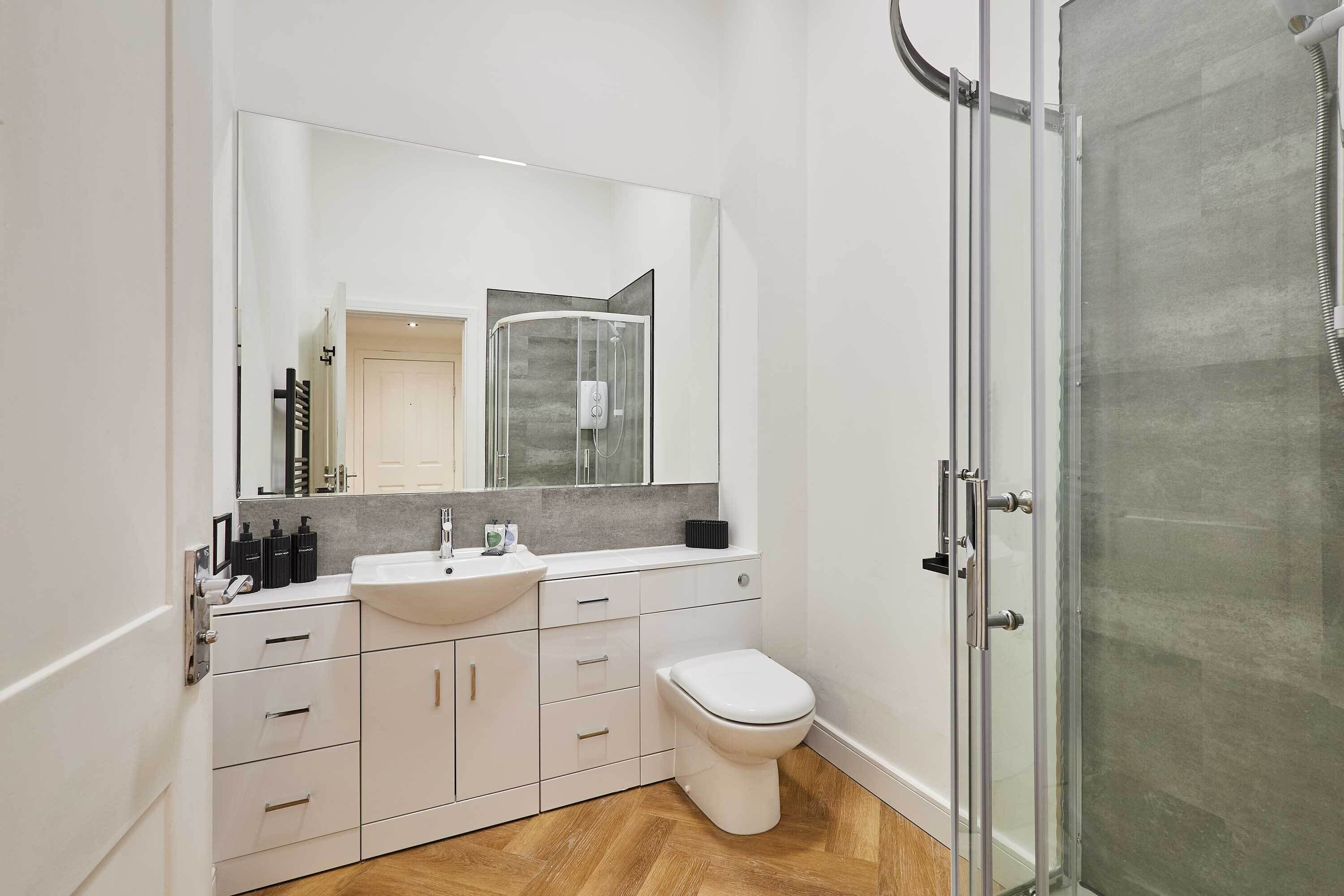 Casa de banho moderna com lavatório branco, espelho e sanita, e área de duche separada por vidro.
