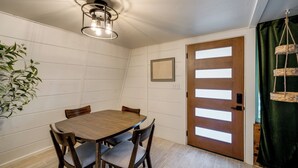 Uma área de refeições com mesa e cadeiras de madeira, luminária pendente, porta com painel de luz e cortina verde.