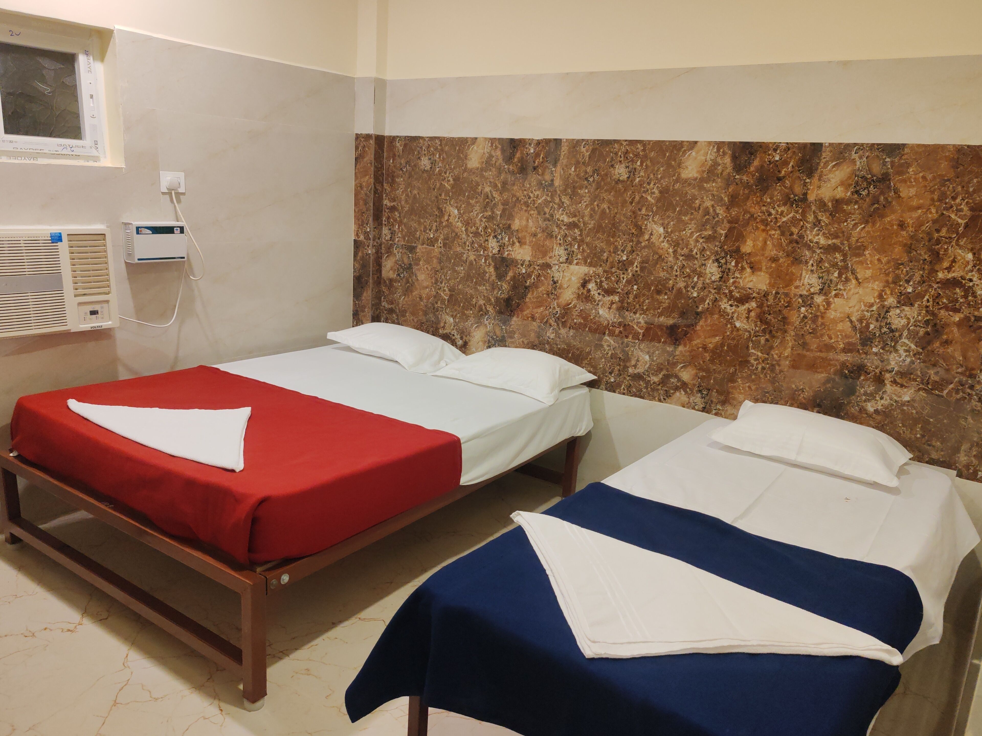 Quarto de hotel com duas camas de solteiro, cada uma com colcha vermelha e branca. Há um ar condicionado fixado na parede à esquerda.