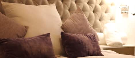 Uma cama com cabeceira estofada, várias almofadas e um edredom branco bem arrumado.