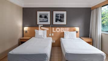 Quarto de hotel com duas camas de solteiro, cabeceira de madeira, mesa de cabeceira com abajur e duas imagens emolduradas na parede.