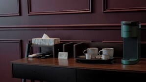 Uma mesa de madeira escura com uma cafeteira Keurig, um suporte com duas chávenas, uma caixa de lenços de papel e um comando à distância.