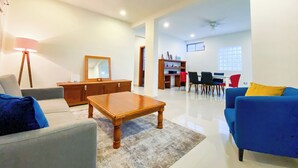 Uma sala de estar espaçosa com uma mesa de centro de madeira, um sofá azul, uma poltrona cinza e uma cadeira vermelha. Há uma estante de livros, um abajur e um ventilador de teto.