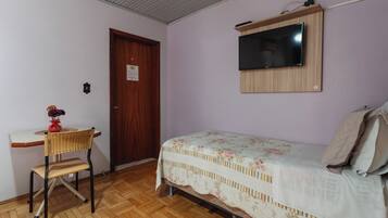 Um quarto com cama, televisão na parede, escrivaninha com cadeira e uma porta.