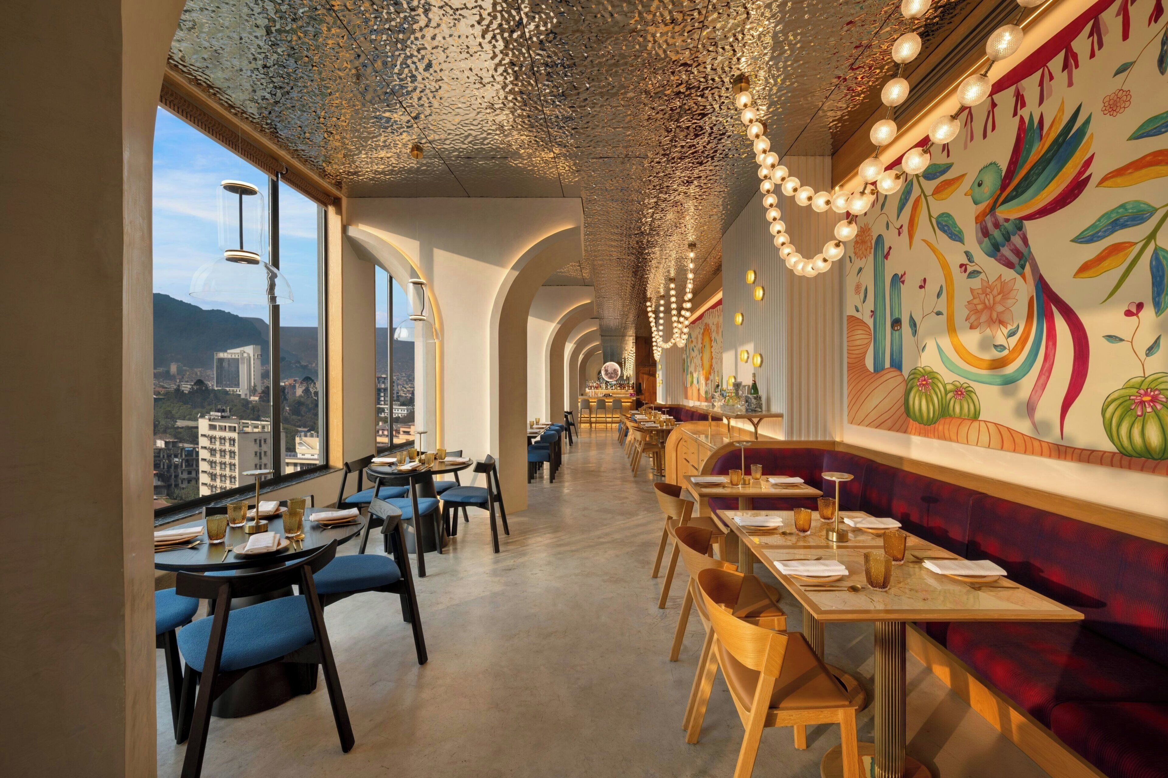 Um restaurante com mesas e cadeiras, um mural na parede e vista para a cidade.