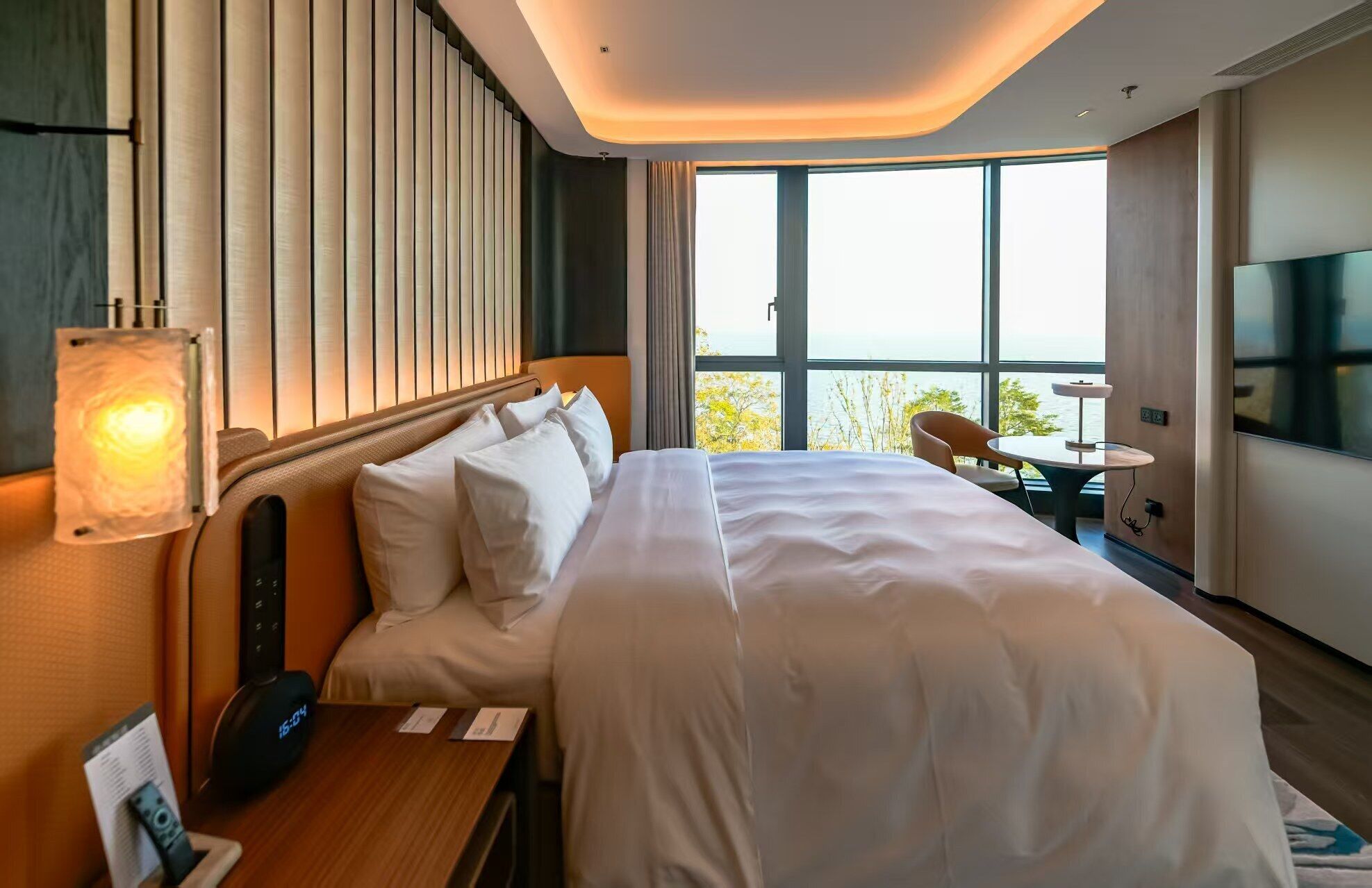 Quarto de hotel com cama, mesa de cabeceira, luminária, televisão e vista para o exterior.