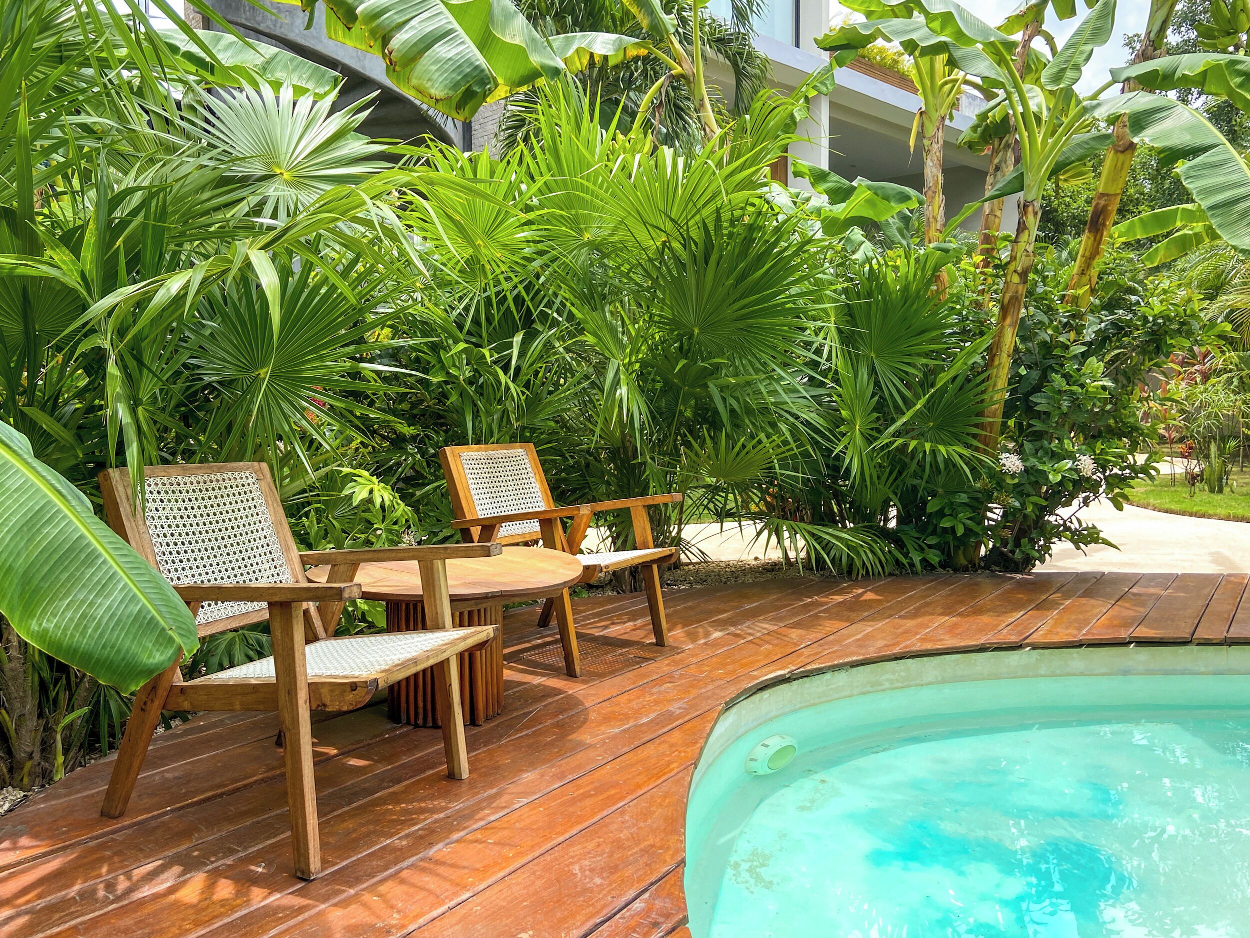 Um deck de madeira com duas cadeiras de vime, cercado por plantas tropicais exuberantes e uma piscina cristalina.