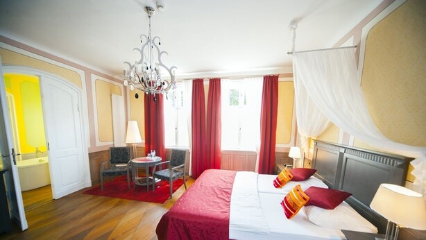 Quarto de hotel com uma cama grande, um candelabro, um espaço de estar com mesa e cadeiras, e vista para o exterior pelas janelas.