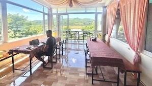 Uma área de refeições com vista para colinas verdes, grandes janelas e uma mesa com cadeiras.