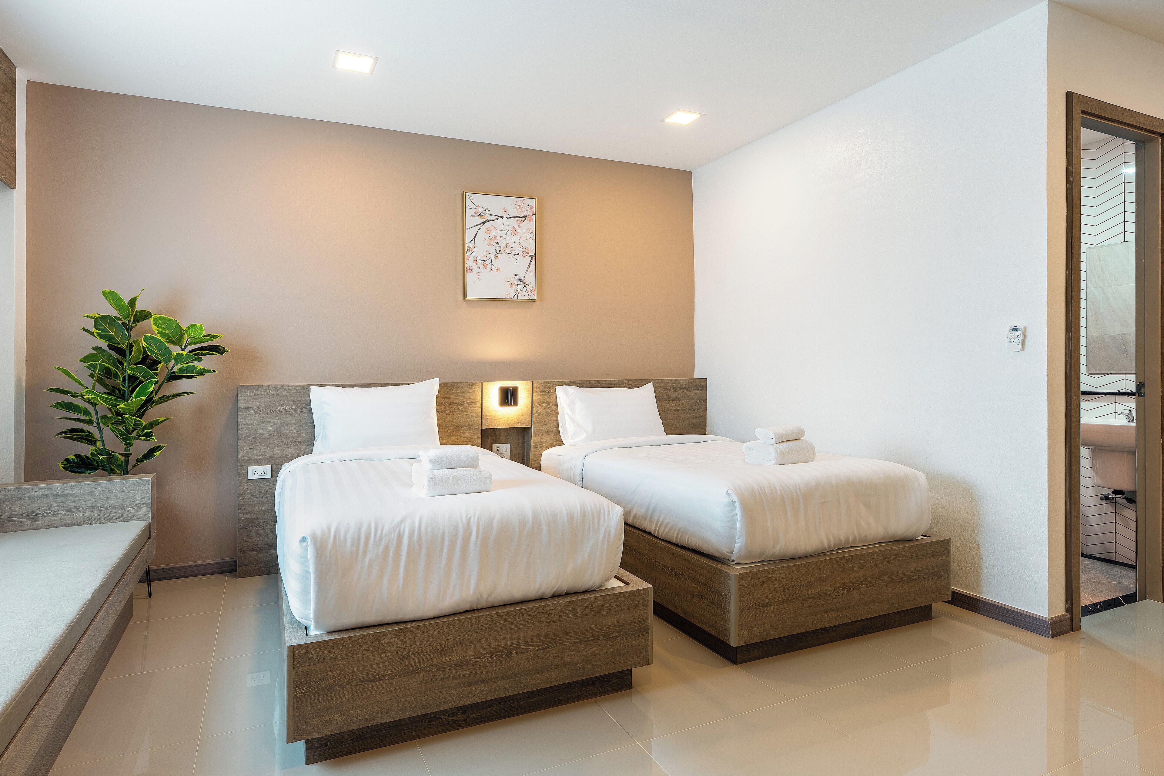Quarto de hotel moderno com duas camas, uma mesinha de cabeceira, um vaso de planta e um banheiro visível por uma porta aberta.