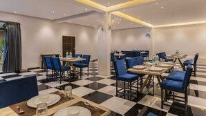 Um espaço de refeições de hotel com chão quadriculado, cadeiras estofadas azuis e mesas preparadas com pratos e copos.